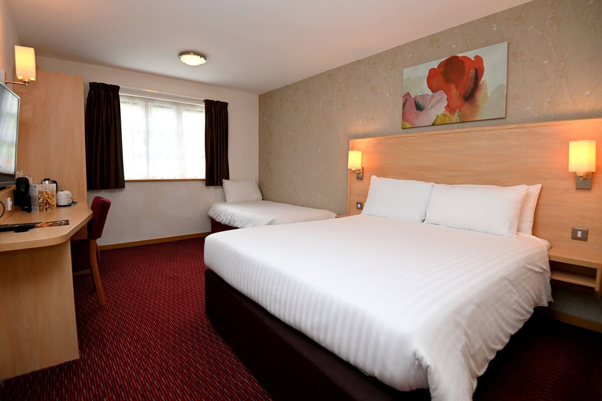 Quarto de hotel com uma cama grande, uma escrivaninha com cadeira, uma janela com cortinas e um quadro na parede.