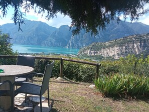 Terraço com cadeiras de vime e uma mesa, com vista para um lago azul-turquesa e montanhas.