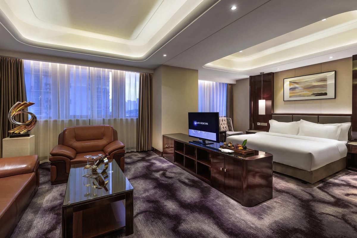 Quarto de hotel com sofá, televisão, cama, mesa de centro e uma janela grande com cortinas.