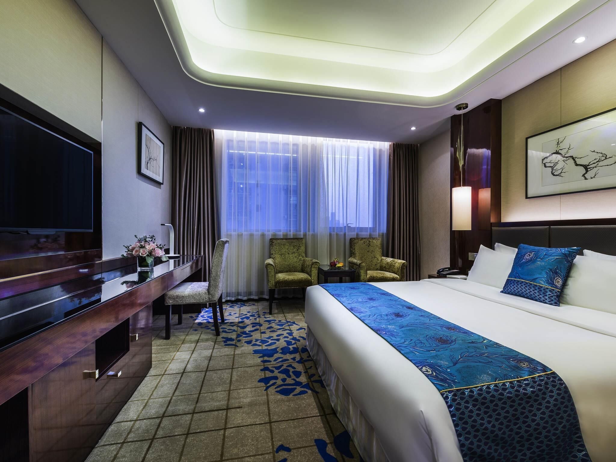 Quarto de hotel com cama, escrivaninha com televisão, duas cadeiras, janela com cortinas e um quadro na parede.