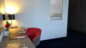 Quarto de hotel com uma escrivaninha de madeira, uma cadeira vermelha, uma luminária e uma janela com persianas.