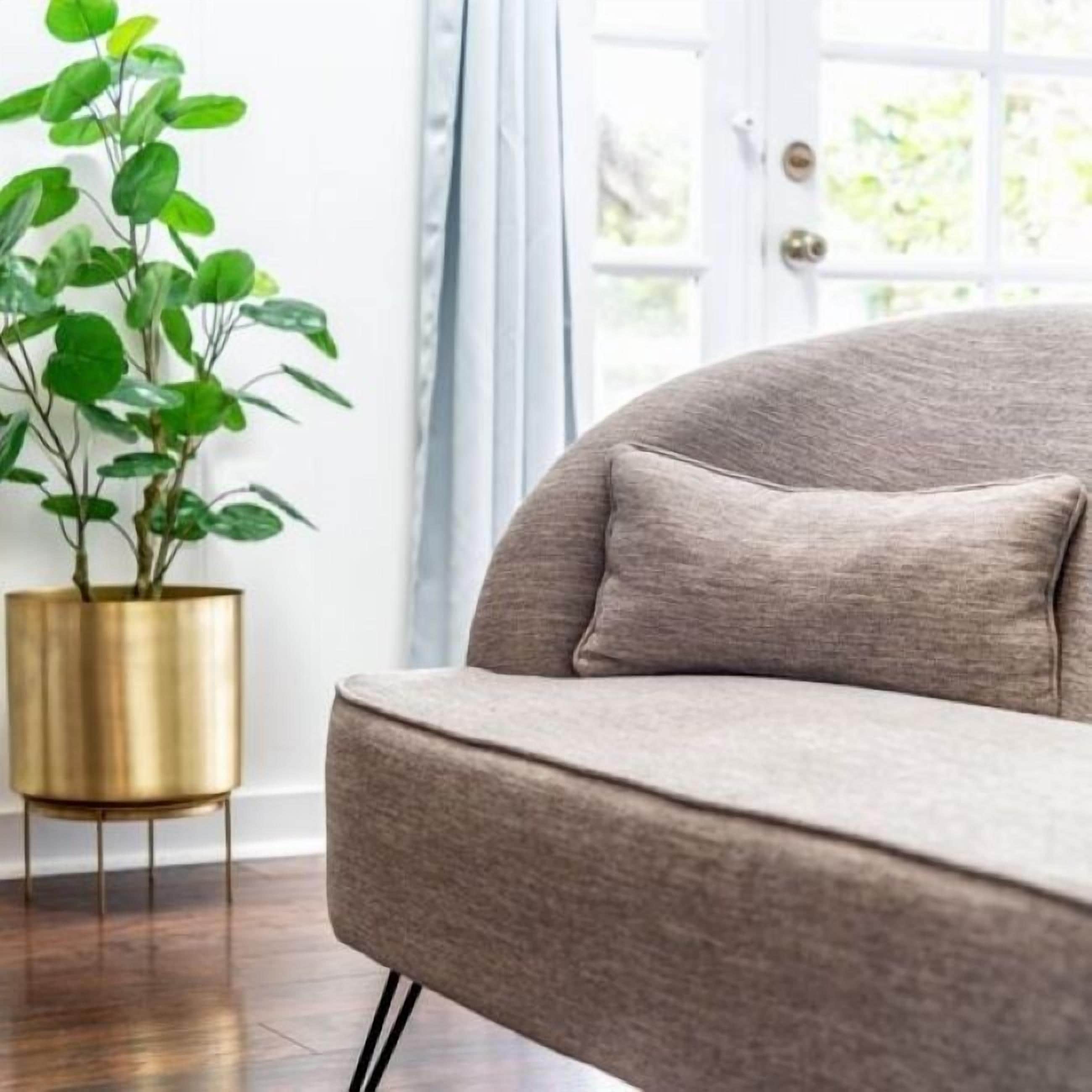 Uma sala de estar moderna com um sofá cinzento, um cachepot dourado com plantas verdes e uma janela com cortinas.