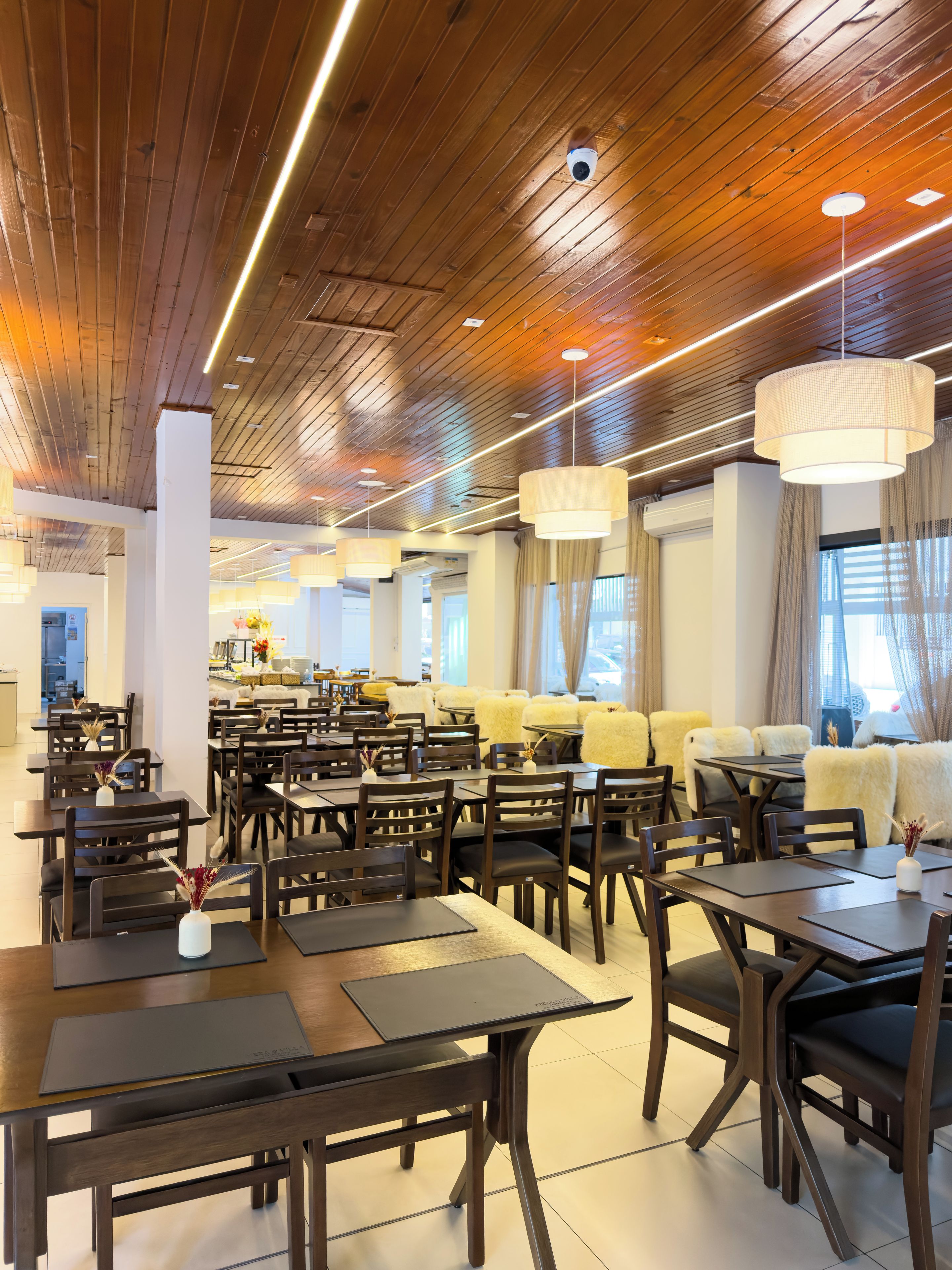 Um restaurante espaçoso com vigas de madeira no teto, iluminação pendente moderna e diferentes opções de assento, incluindo mesas e sofás acolchoados.