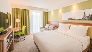 Quarto de hotel com cama, escrivaninha, cadeira, luminária e uma janela grande com cortinas.