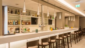 Uma área de bar com um balcão comprido, várias prateleiras com garrafas, copos e alguns banquetas.
