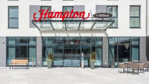 Entrada de um hotel Hampton by Hilton, com fachada de vidro, um banco de madeira e área de estar externa.