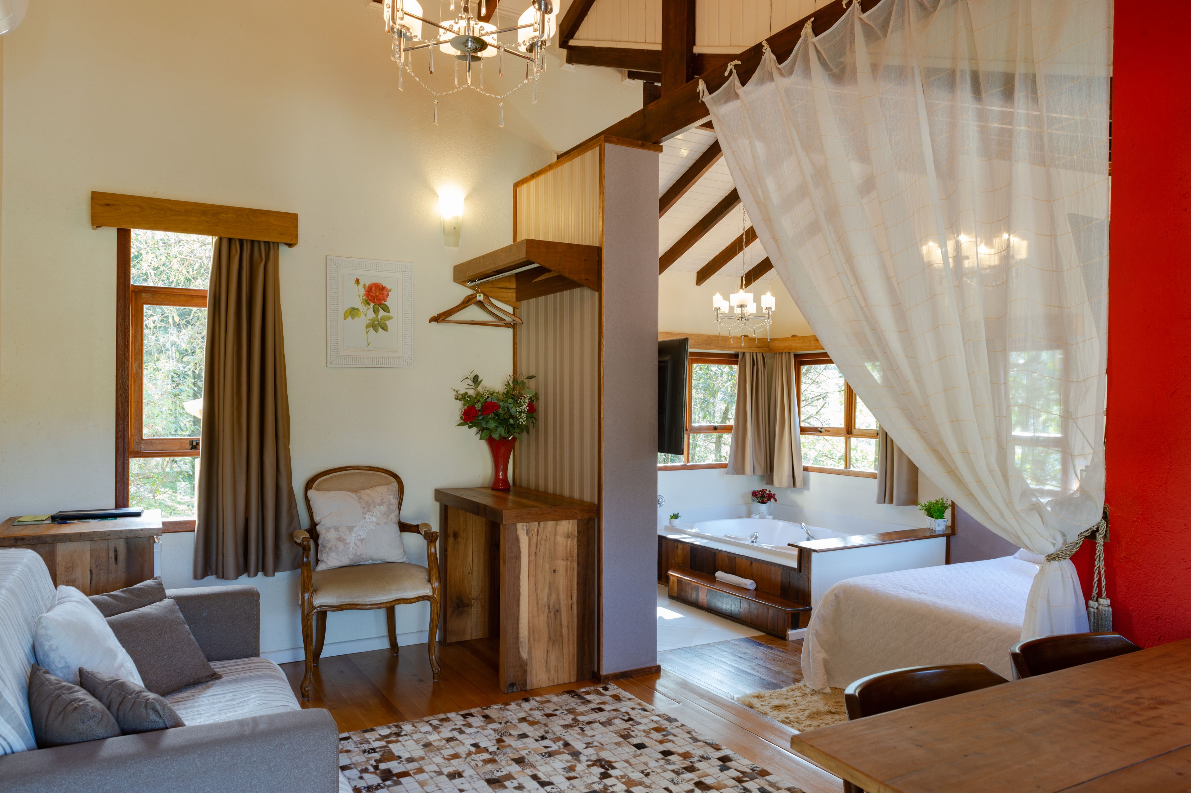 Quarto com sofá, poltrona, mesa de madeira e cama. Há uma janela com cortinas, um candelabro e uma planta sobre a mesa.