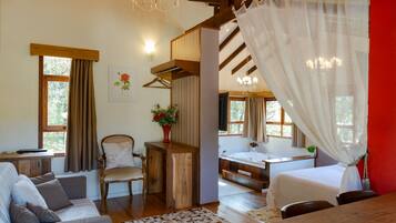 Quarto com sofá, poltrona, mesa de madeira e cama. Há uma janela com cortinas, um candelabro e uma planta sobre a mesa.