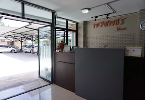 Uma área de recepção com porta de vidro, balcão de recepção, relógio e placa com a inscrição 'MAVIMOS Toma'.