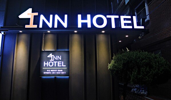 Entrada de hotel com uma placa chamativa exibindo o nome "4INN HOTEL" e uma placa menor com informações de contato.