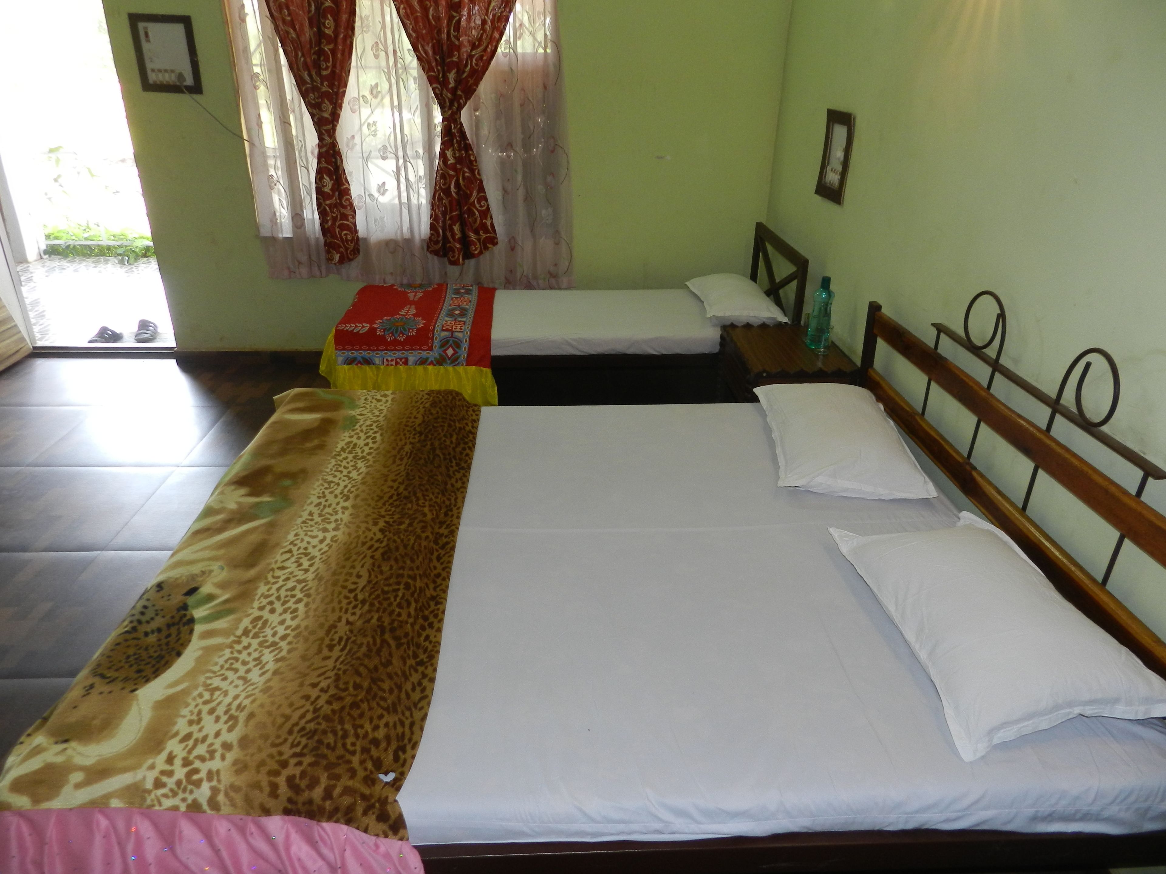 Quarto com duas camas, uma com colcha de onça e outra com roupa de cama branca. Há uma mesinha com toalha vermelha, uma garrafa verde e um quadro na parede.