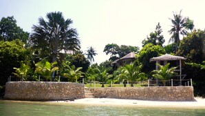 Um resort à beira-mar com um muro de pedra, palmeiras e uma construção com telhado de palha.