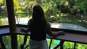 Uma pessoa sentada à mesa de madeira com vista para um jardim exuberante e uma piscina.