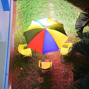 Um guarda-chuva colorido, duas cadeiras amarelas e uma pequena mesa numa área gramada.