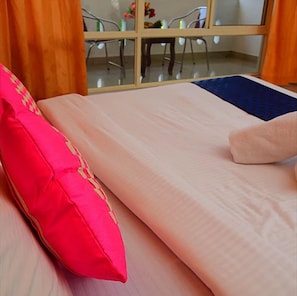 Uma cama com um travesseiro rosa e um travesseiro azul, uma cabeceira de madeira e vista para uma varanda com cadeiras e flores.