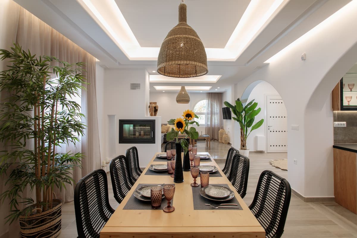 Uma sala de jantar com uma mesa comprida posta para seis pessoas, rodeada por cadeiras pretas, uma grande planta em vaso e um pendente tecido.