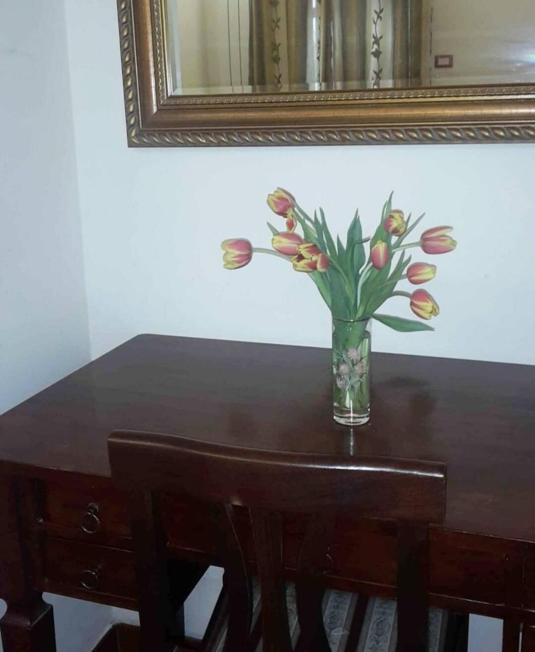 Uma mesa de jantar com um vaso de tulipas, um espelho na parede e cadeiras de madeira.