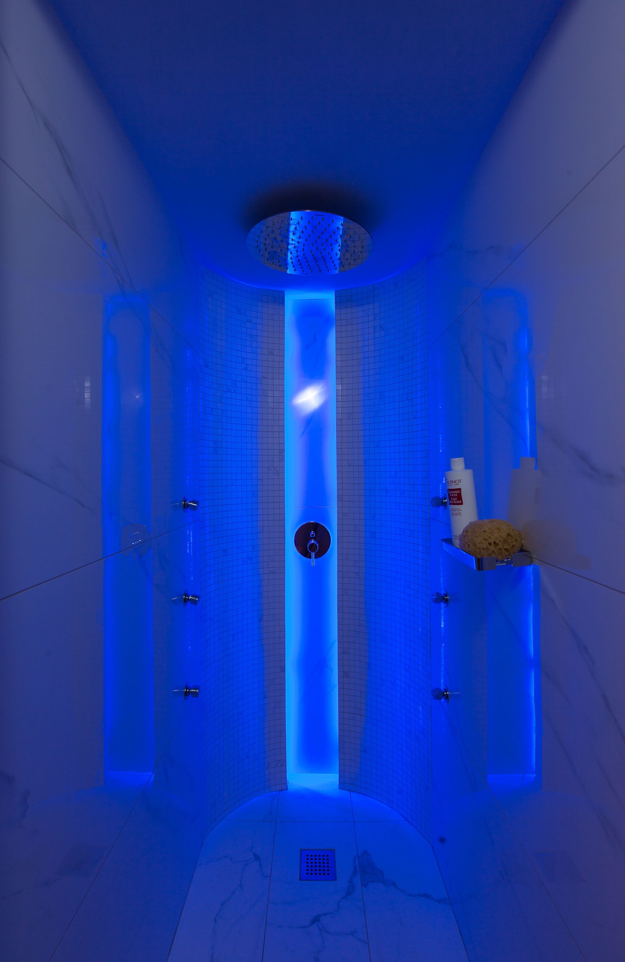 Casa de banho com chuveiro iluminado em azul, um porta-toalhas e uma garrafa de champô.