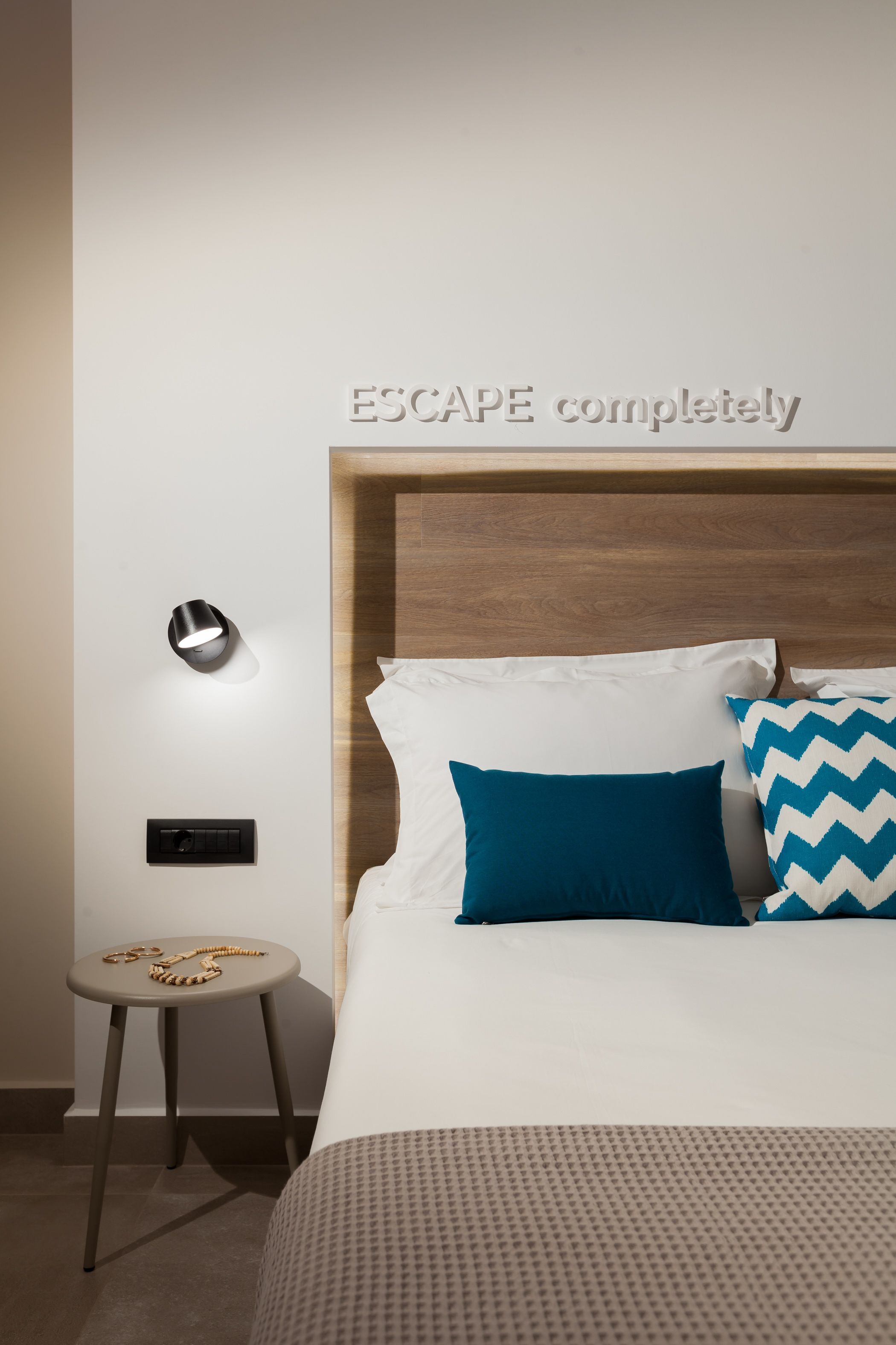 Cama bem arrumada com cabeceira, abajur e mesinha redonda com objeto decorativo.