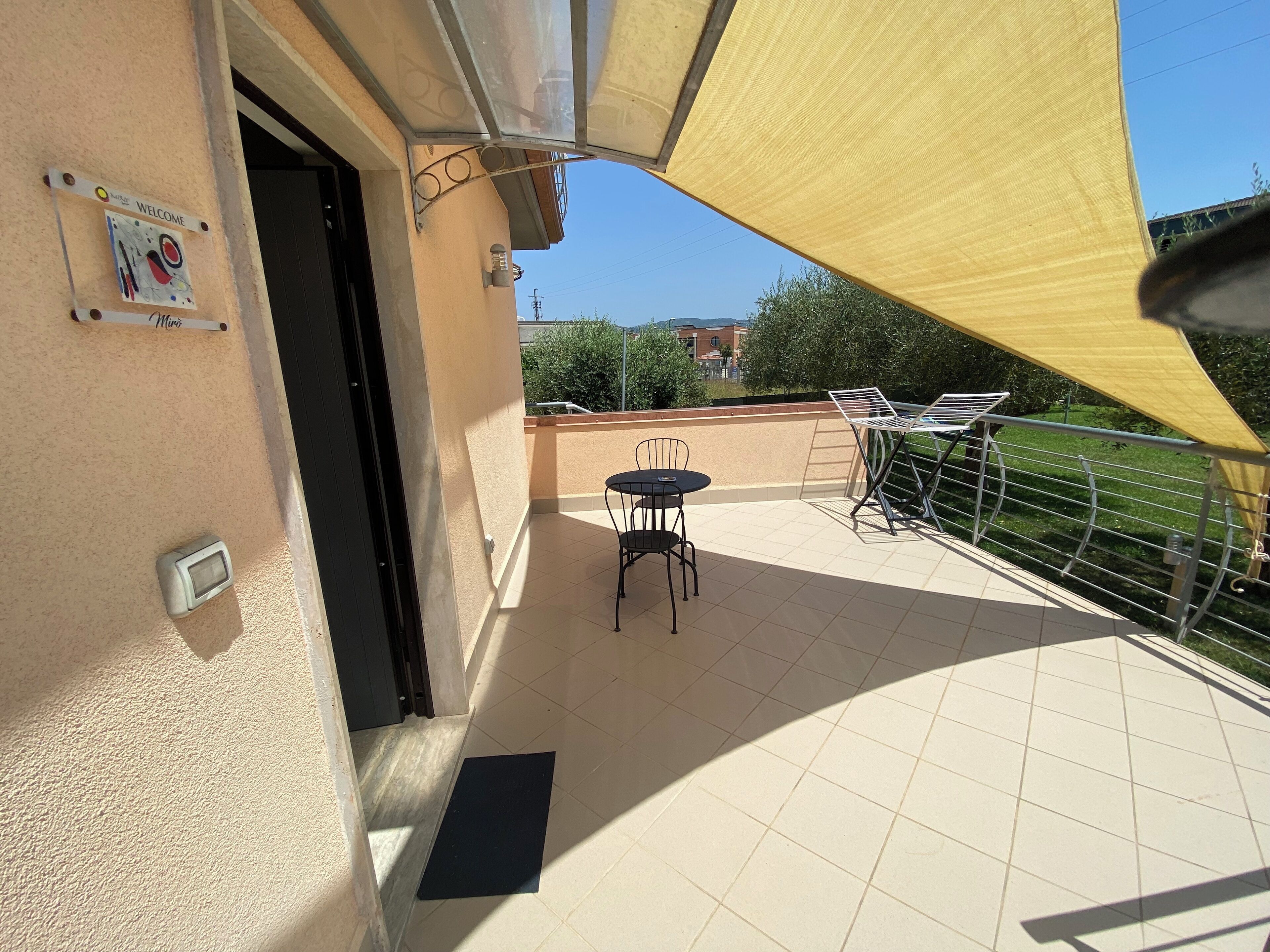 Uma varanda com mesa e cadeiras, um toldo amarelo e uma placa de boas-vindas.