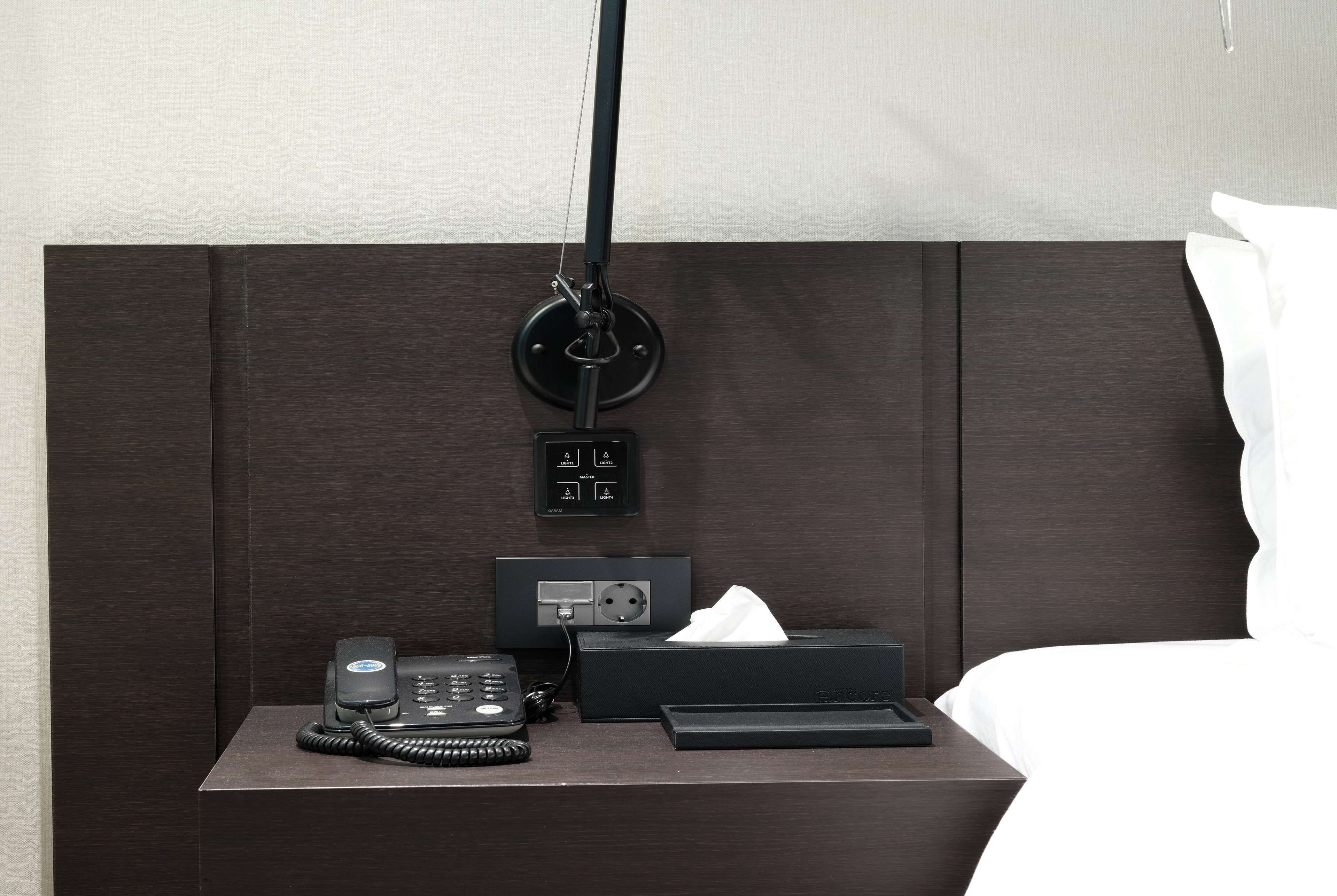 Quarto de hotel bem arrumado, com uma mesa de cabeceira que possui um telefone, caixa de lenços e tomada.