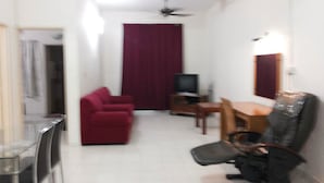 Um espaço de estar compacto, com um sofá vermelho, uma televisão sobre um suporte de madeira, uma mesa de jantar com cadeiras e um ventilador de teto.