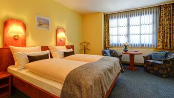 Quarto de hotel com uma cama grande, duas luminárias de cabeceira, uma mesa pequena com um vaso, duas poltronas e uma janela com cortinas.