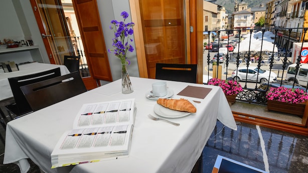 Uma mesa posta com um menu, um croissant e um vaso de flores roxas num restaurante com vista para uma rua movimentada e edifícios.