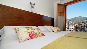 Quarto de hotel com cabeceira de madeira, duas camas com almofadas florais, vista da cidade através de uma varanda e um vaso com flores na parede.