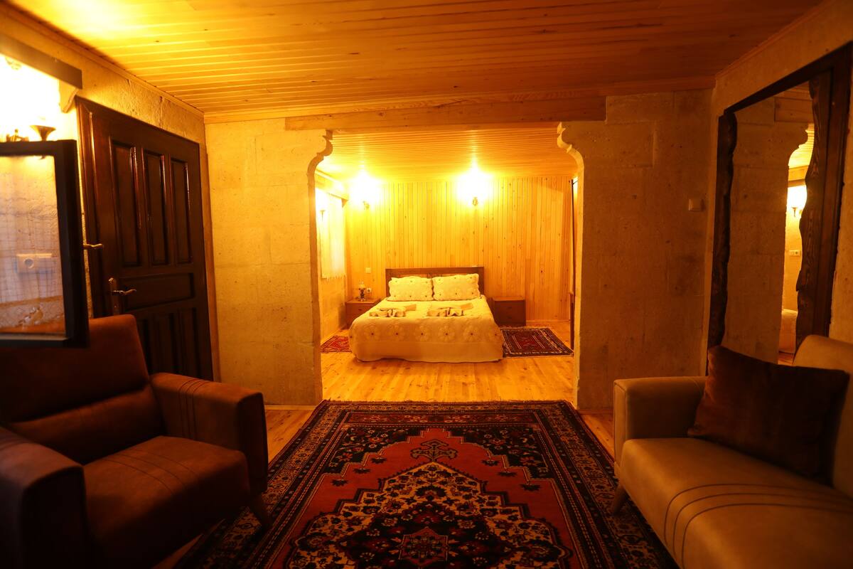Quarto com piso de madeira, cama com roupa de cama branca, duas poltronas e um tapete estampado.