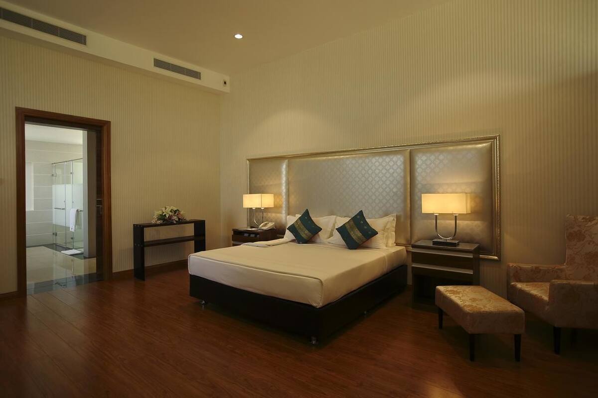 Quarto de hotel com uma cama grande, duas mesas de cabeceira com luminárias, uma cadeira e uma pequena mesa com um vaso.
