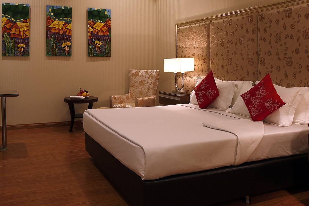 Quarto de hotel com uma cama grande, uma cadeira, uma mesa pequena com frutas e três quadros na parede.