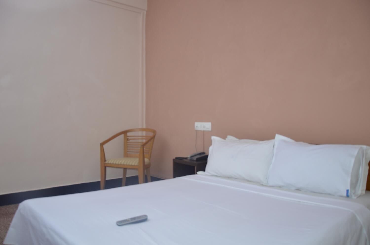 Uma cama de solteiro com roupa de cama branca, uma cadeira de madeira, uma mesa de cabeceira com abajur e uma tomada na parede.