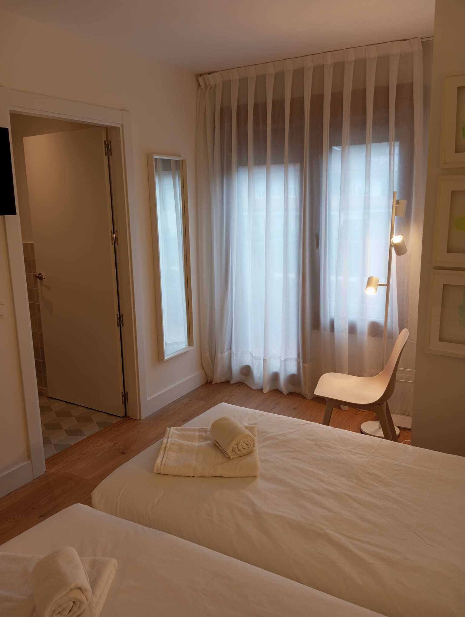Quarto de hotel com cama, cadeira, escrivaninha e janela com cortinas.