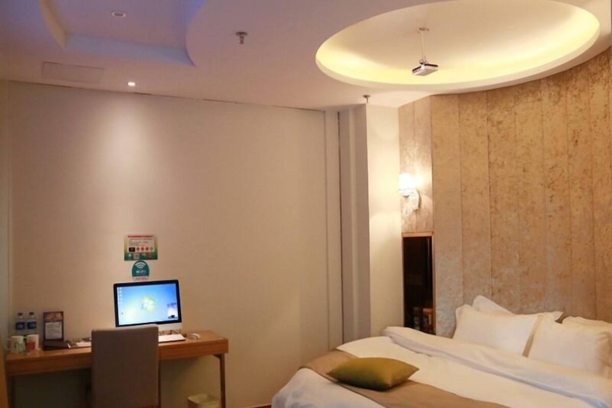 Quarto de hotel com cama, secretária com computador e luminária fixada na parede.