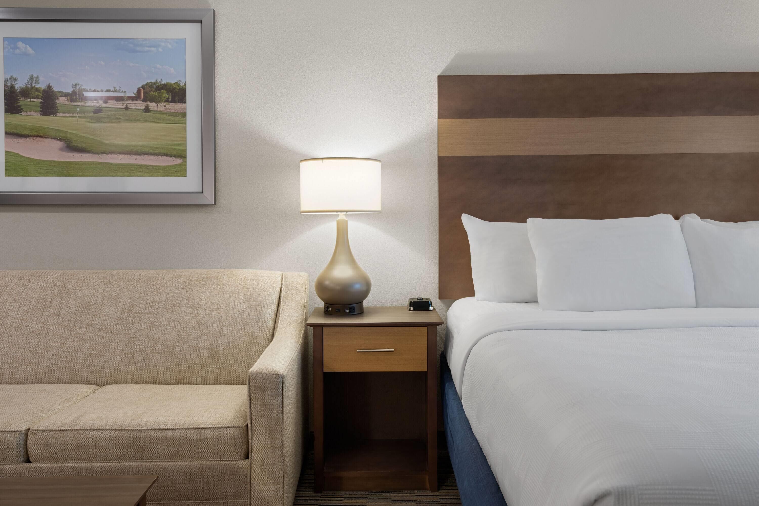 Quarto de hotel com cama, mesa de cabeceira com abajur, quadro com paisagem de campo de golfe e sofá.