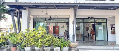 Entrada de hotel com varanda coberta, plantas em vasos e interior visível através de portas de vidro.