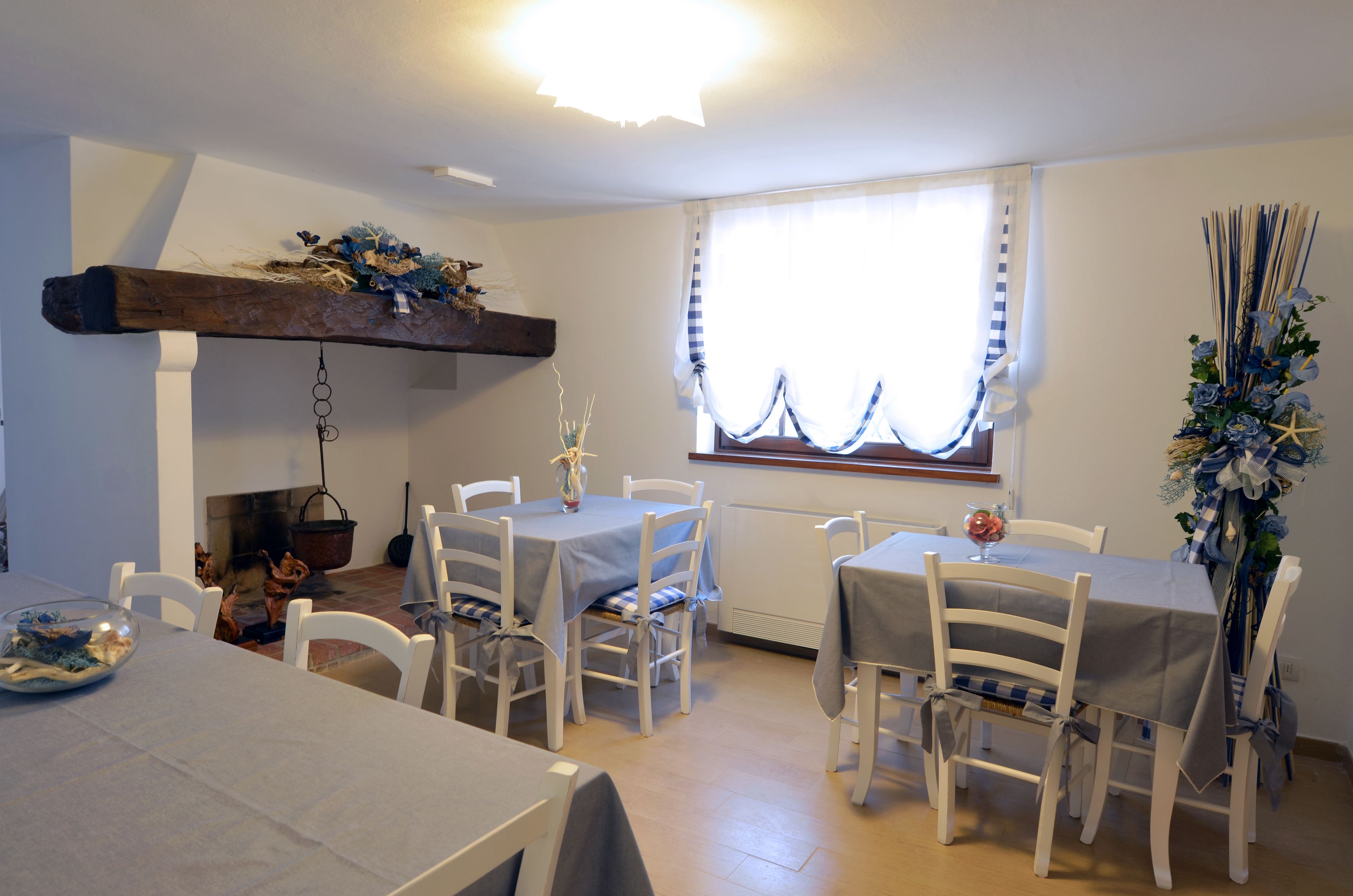 Uma área de refeições com cadeiras e mesas brancas, uma estante de madeira com decorações, uma janela com cortinas e um vaso com flores.