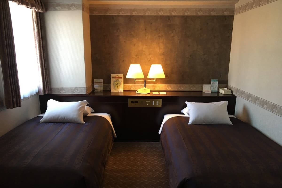 Quarto de hotel com duas camas de solteiro, mesa de cabeceira com luminária, espelho e pequena mesa com telefone e caixa de lenços.