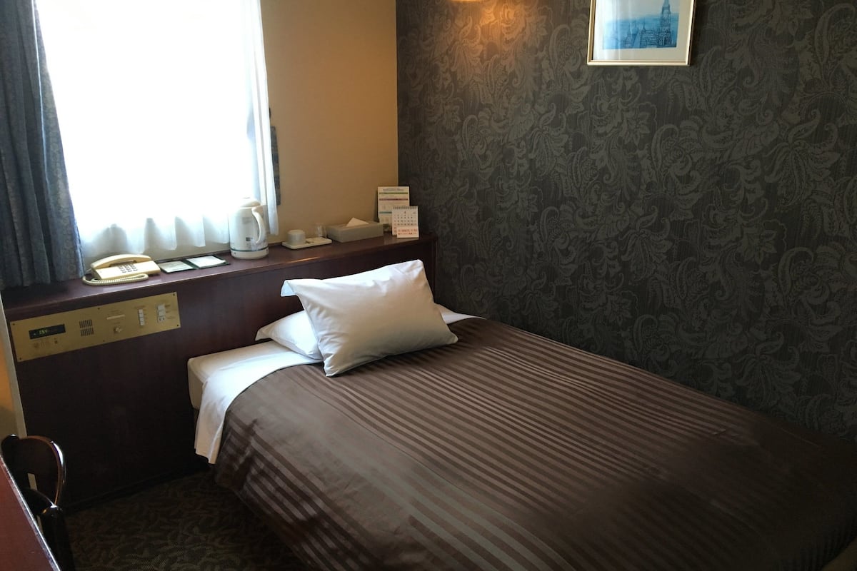 Quarto de hotel com cama, mesa de cabeceira, telefone, relógio e janela com cortinas.