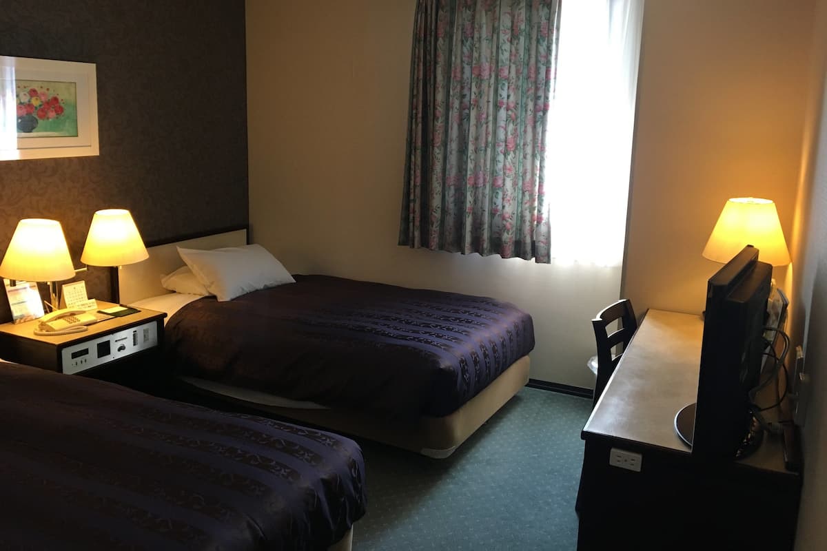 Quarto de hotel com duas camas de solteiro, uma escrivaninha com abajur, telefone e televisão.