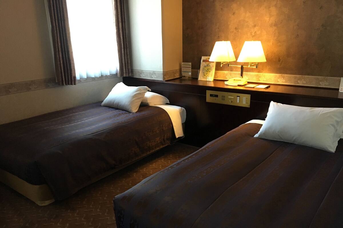 Quarto de hotel com duas camas de solteiro, mesa de cabeceira com abajur e janela com cortinas.