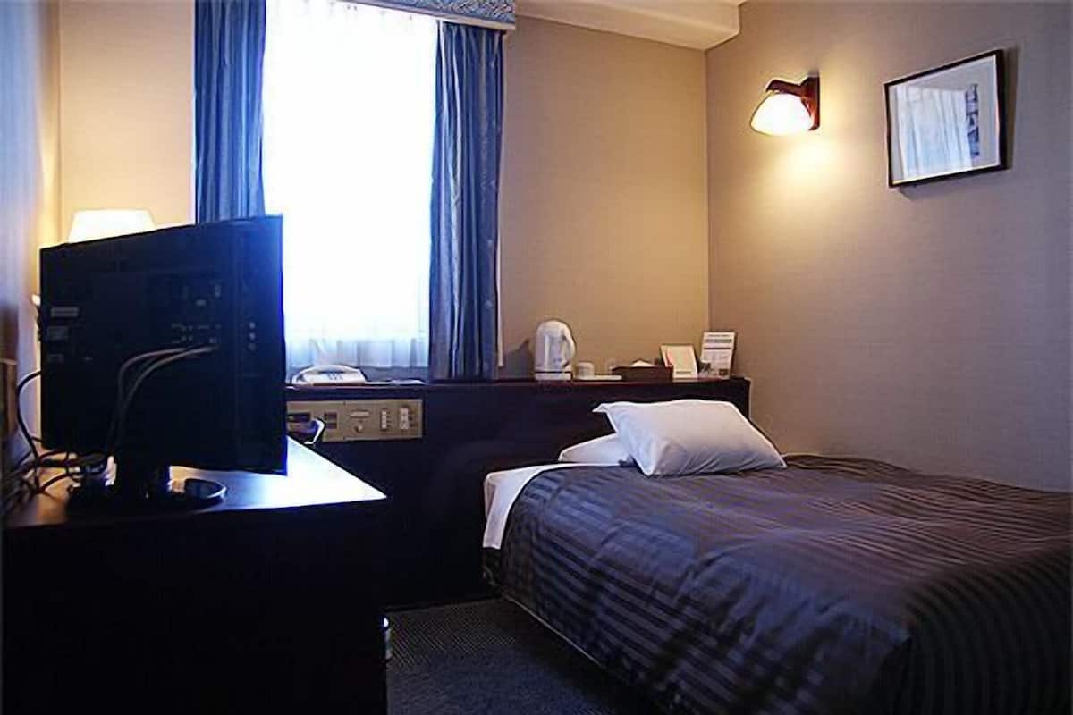 Quarto de hotel com escrivaninha, cama, janela com cortinas, luminária fixada na parede e um quadro emoldurado.