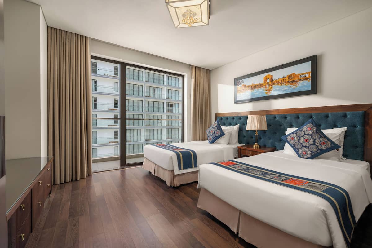 Quarto de hotel com duas camas, uma janela grande com cortinas, um quadro na parede e piso de madeira.
