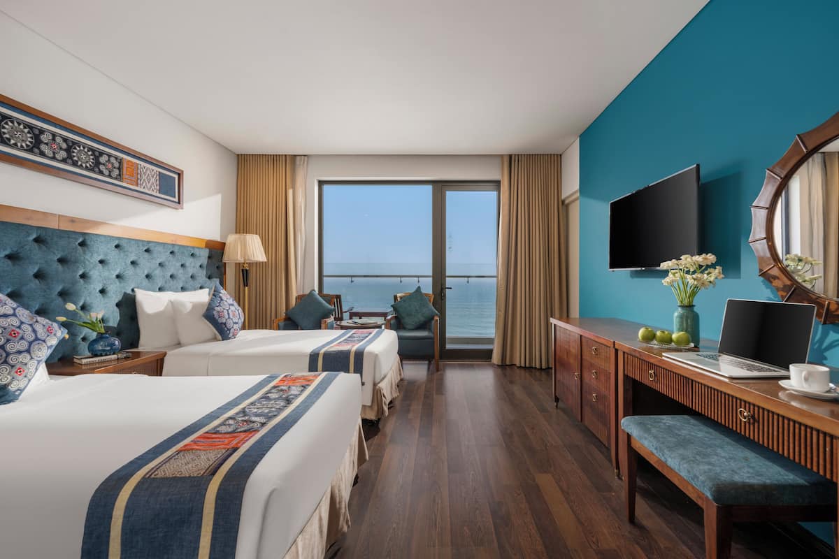 Quarto de hotel com duas camas, uma escrivaninha com um laptop, televisão e vista para o mar.