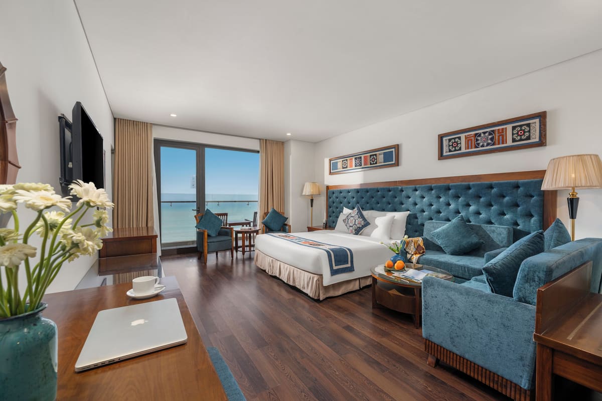 Quarto de hotel com cama, sofá, escrivaninha com laptop, um vaso de flores e vista para o mar.