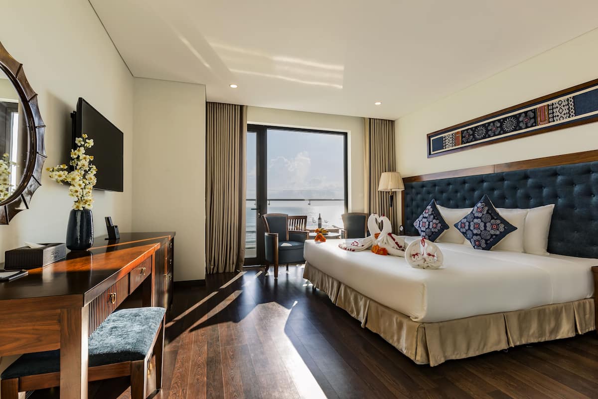 Quarto de hotel com uma cama grande, uma escrivaninha, uma cadeira, uma TV e vista para o mar.