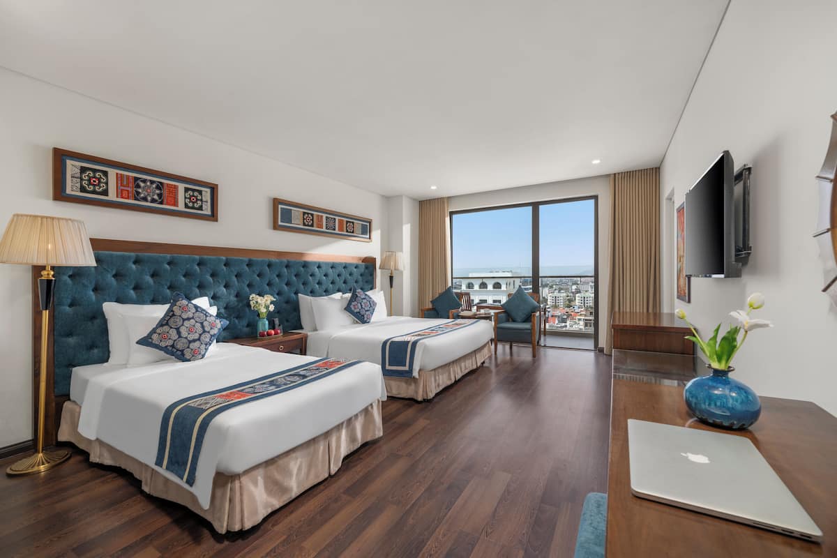 Quarto de hotel com duas camas, uma escrivaninha com um laptop, uma televisão e uma grande janela com vista para a cidade.