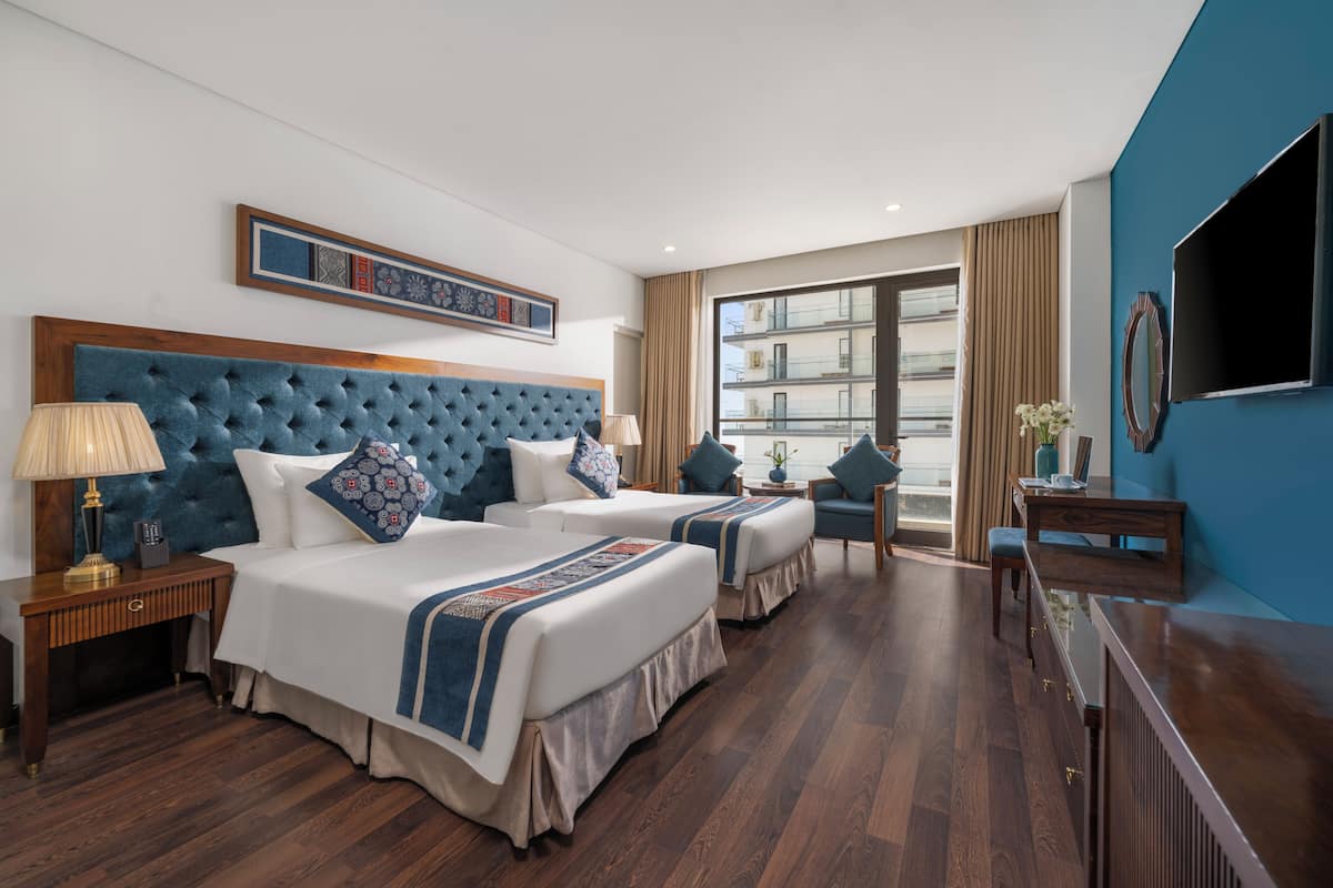Quarto de hotel com uma cama grande, duas mesas de cabeceira, uma TV e uma janela com vista para edifícios.
