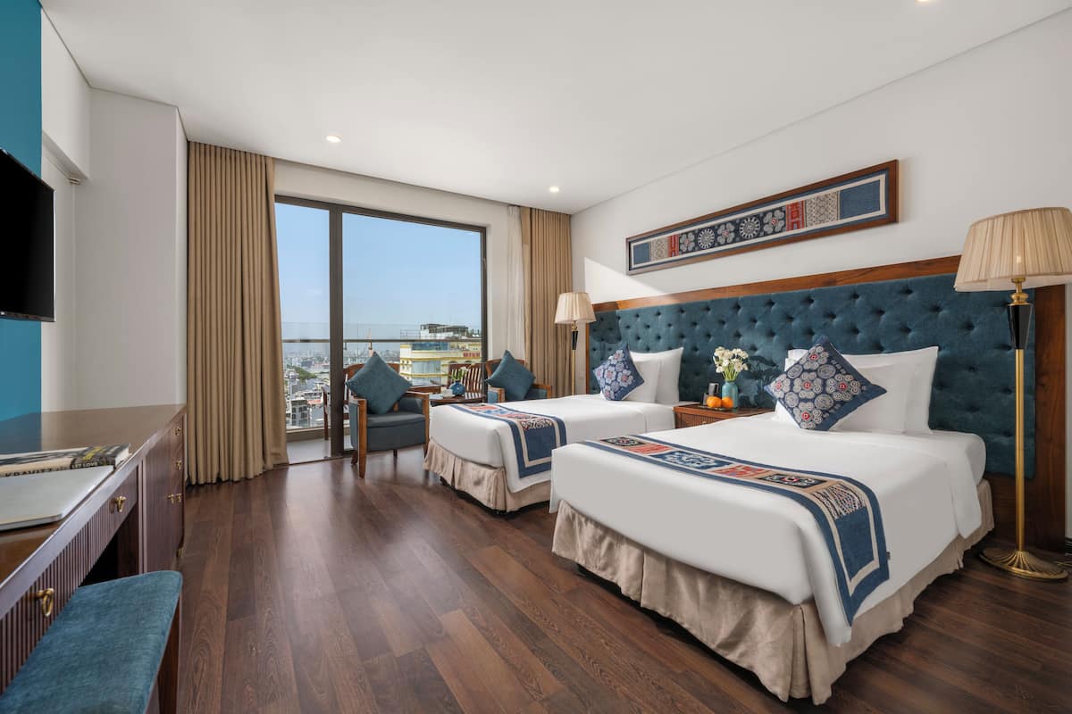 Quarto de hotel com duas camas, uma escrivaninha, uma cadeira, televisão e uma janela grande com vista para a cidade.